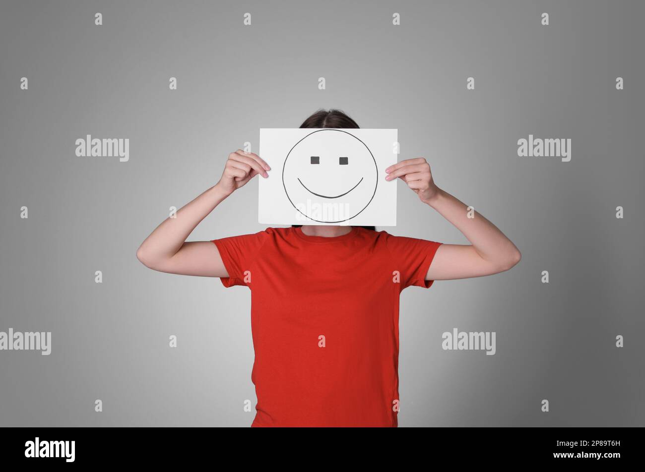 Woman hiding behind sheet of paper with happy face on grey background ...