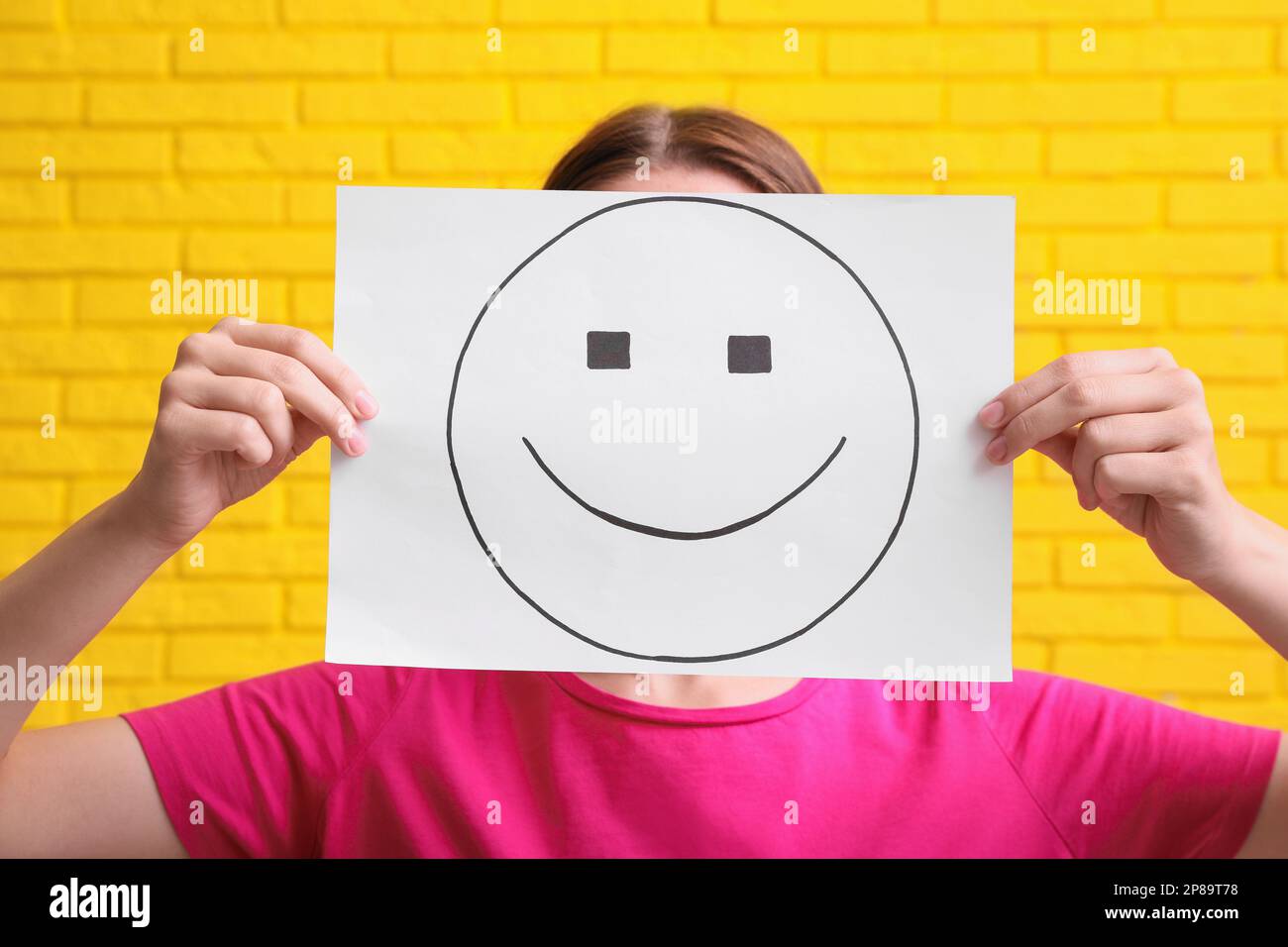 Woman hiding behind sheet of paper with happy face against yellow brick ...