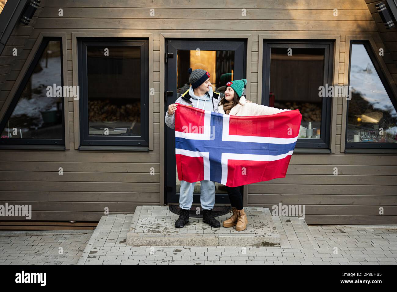 Portrait of couple outside cabin house holding Norway flag ...