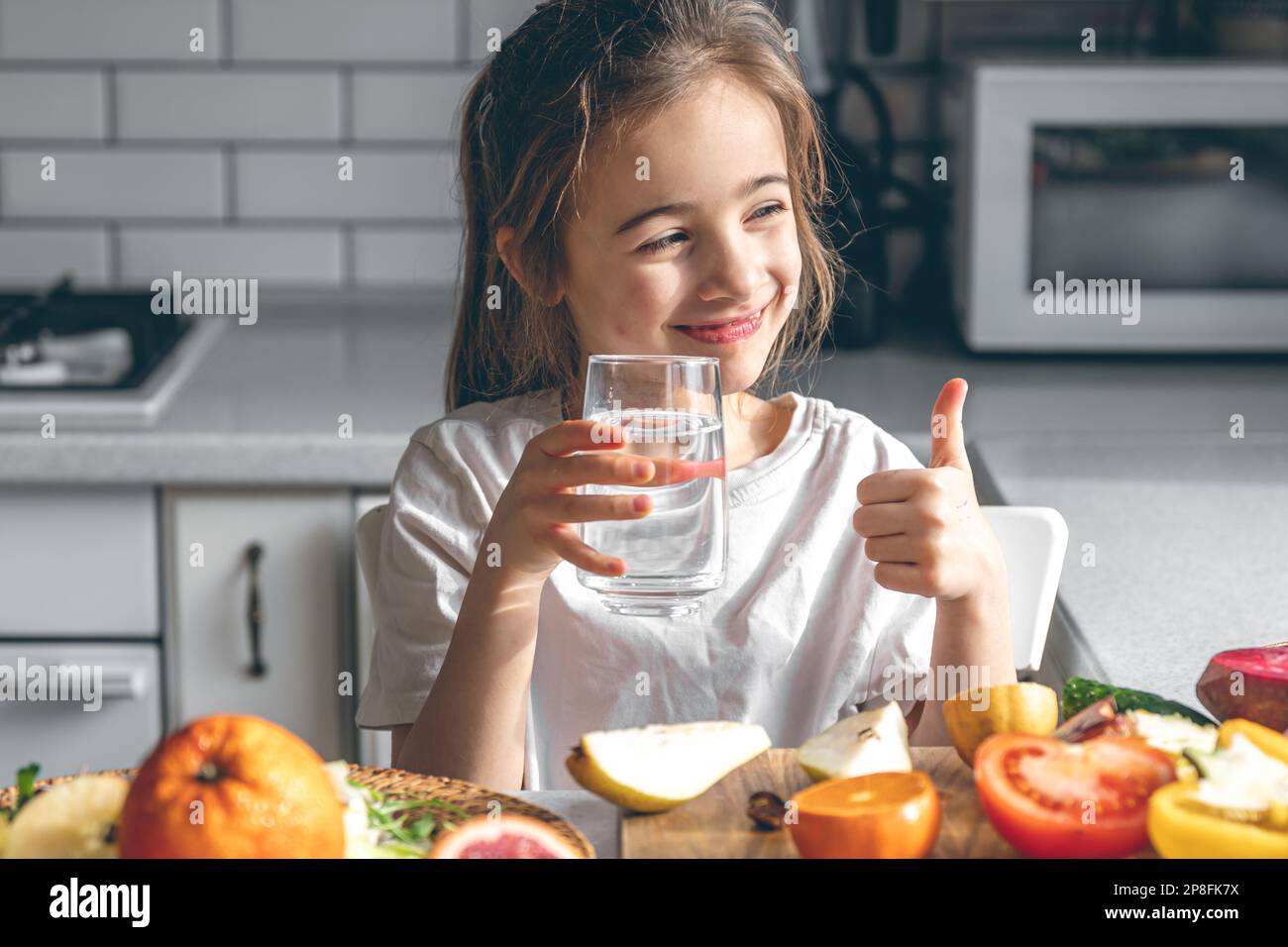 Little girl with a glass of water in a kitchen, with fruits and ...
