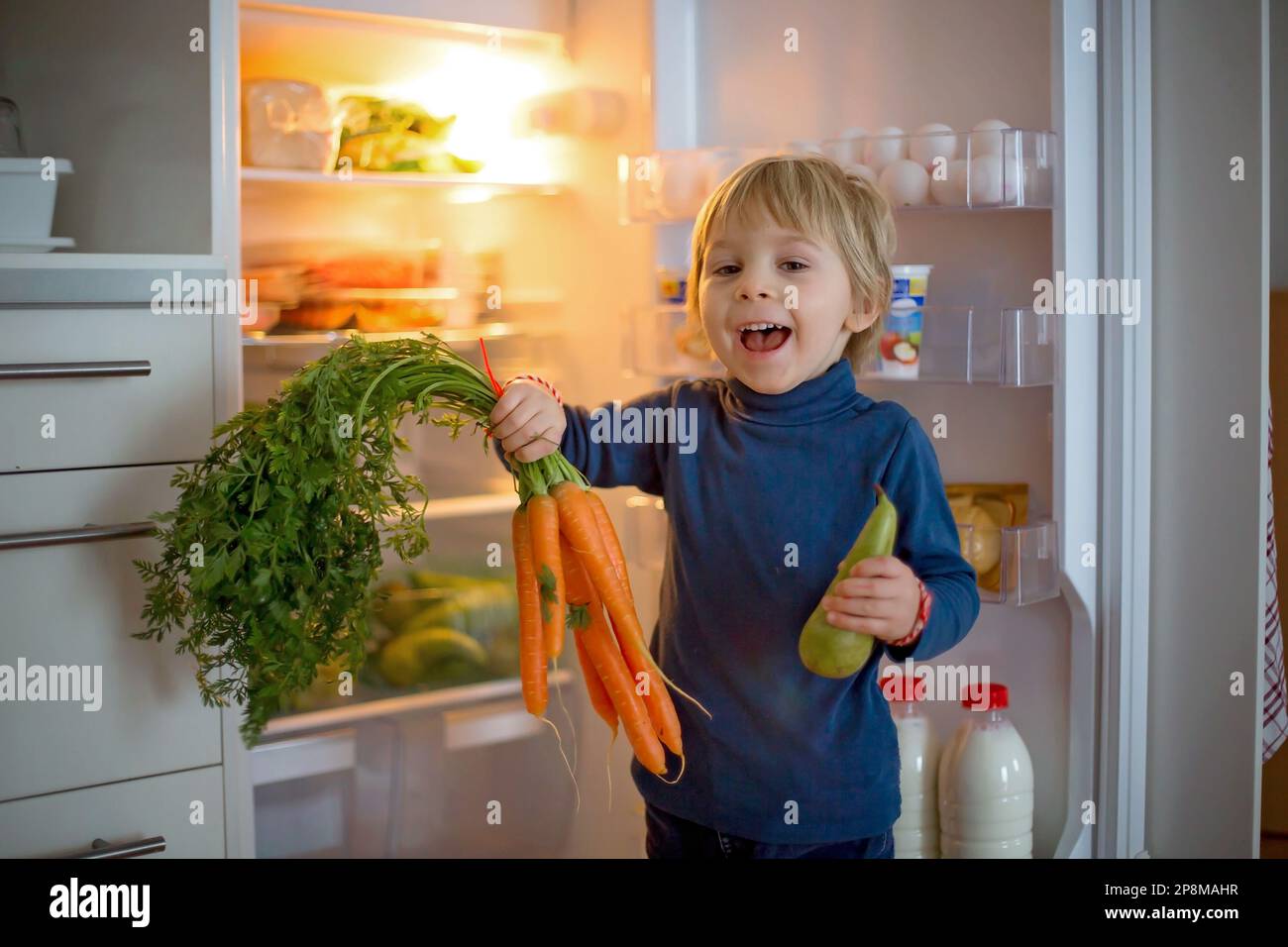 Cute toddler blond child, opening the fridge door and taking fruits adn ...