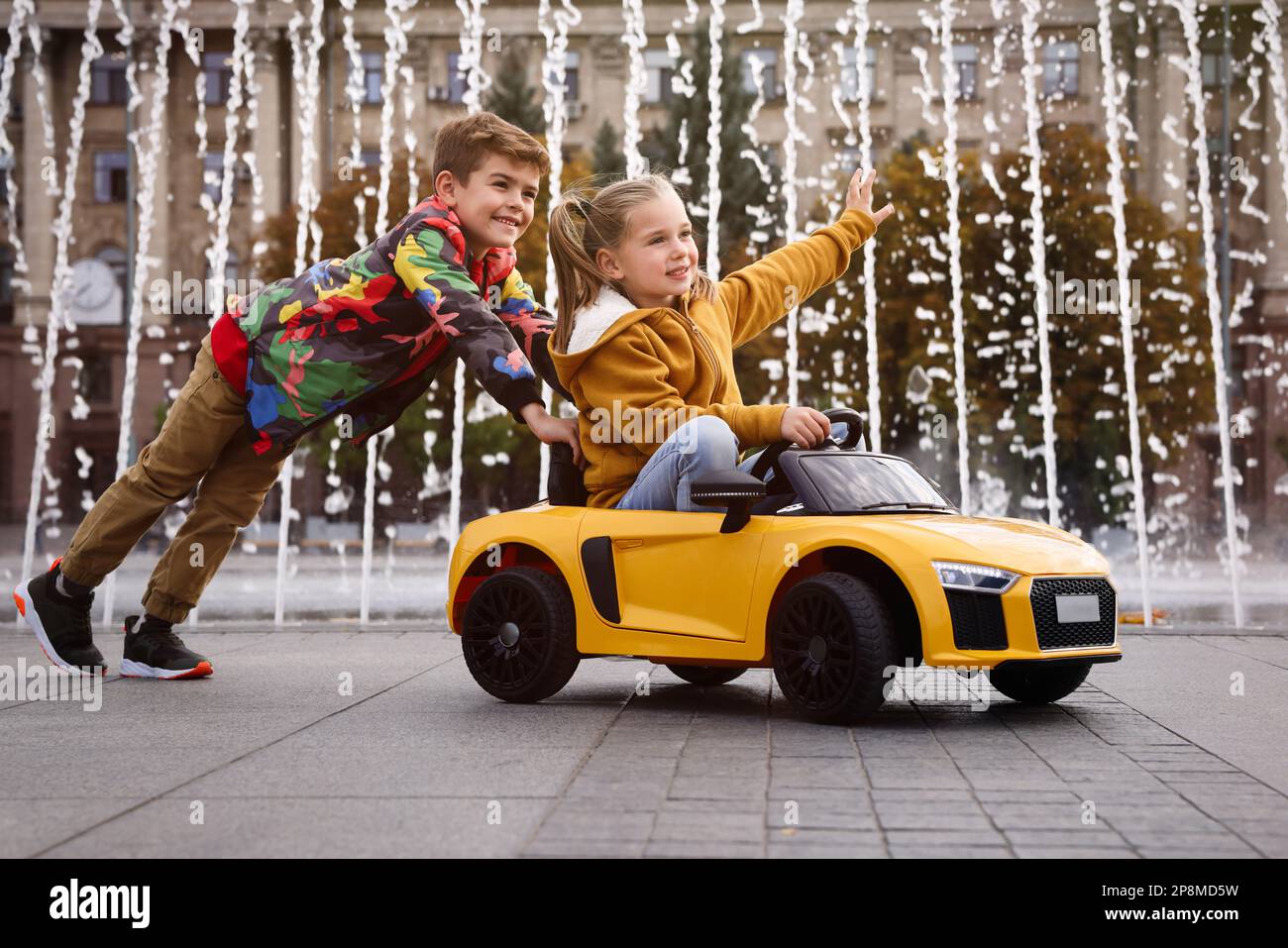 Cute boy pushing children's car with little girl near fountain on city ...