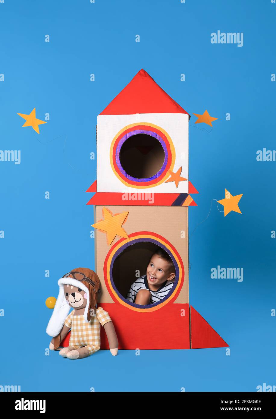 Cute little boy playing with cardboard rocket on light blue background ...
