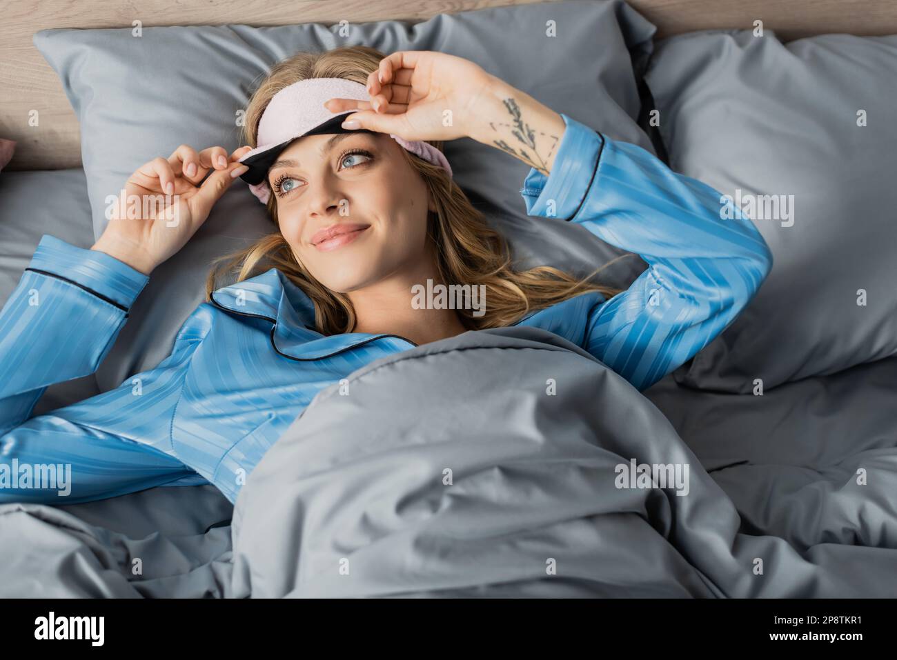 overhead view of tattooed and happy woman in silk pajama adjusting ...