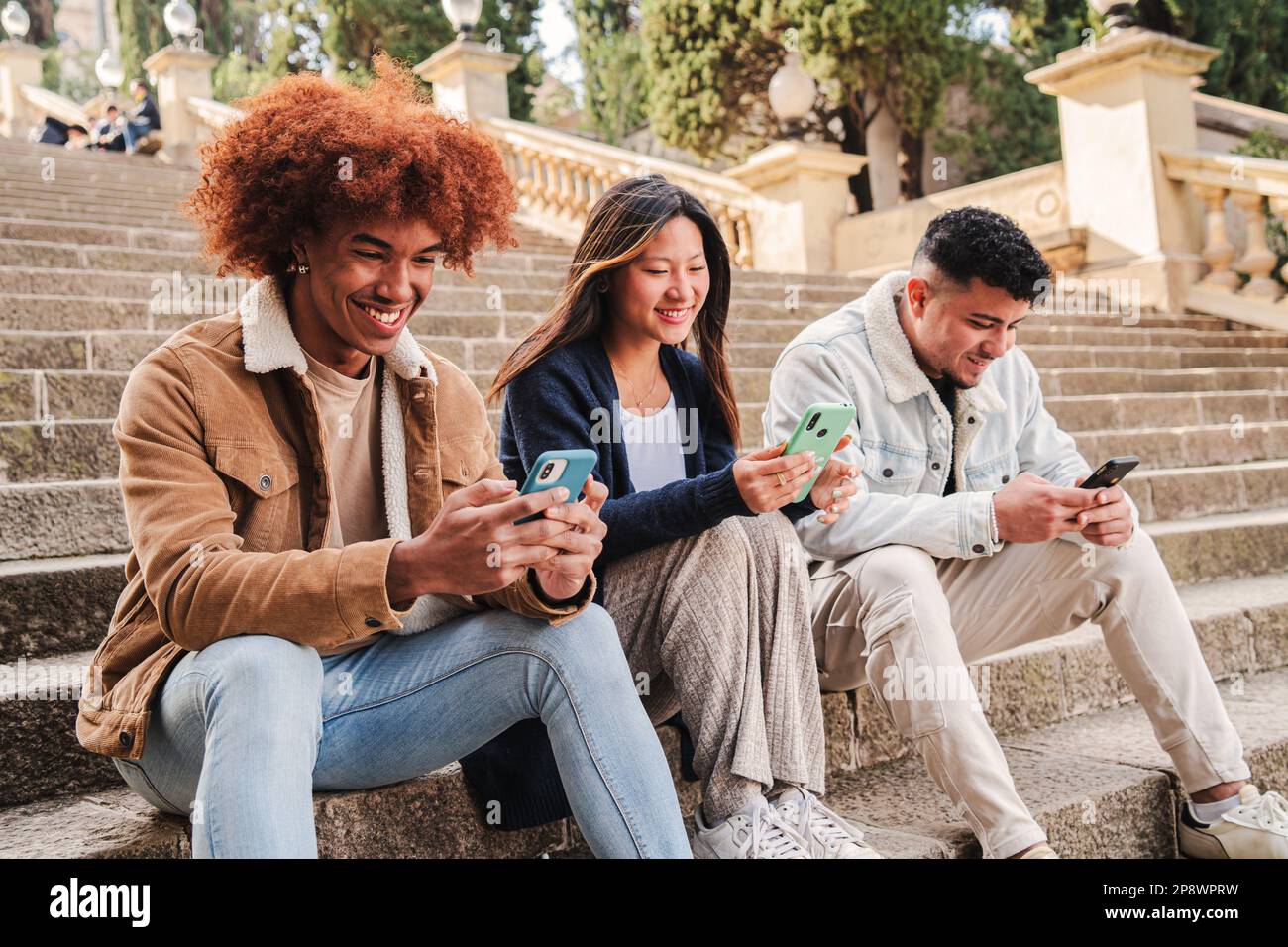Multiracial group young friends enjoying and smiling using their mobile phone app sitting at ...