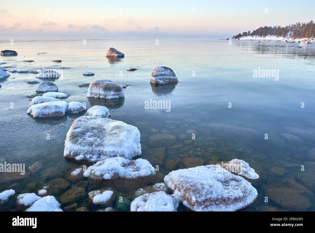 A tranquil shoreline in winter, with snow-covered rocks and a partially ...