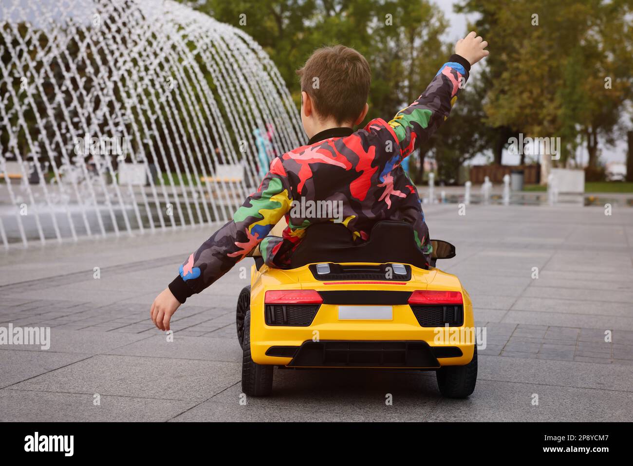Cute little boy driving children's car near fountain on city street ...