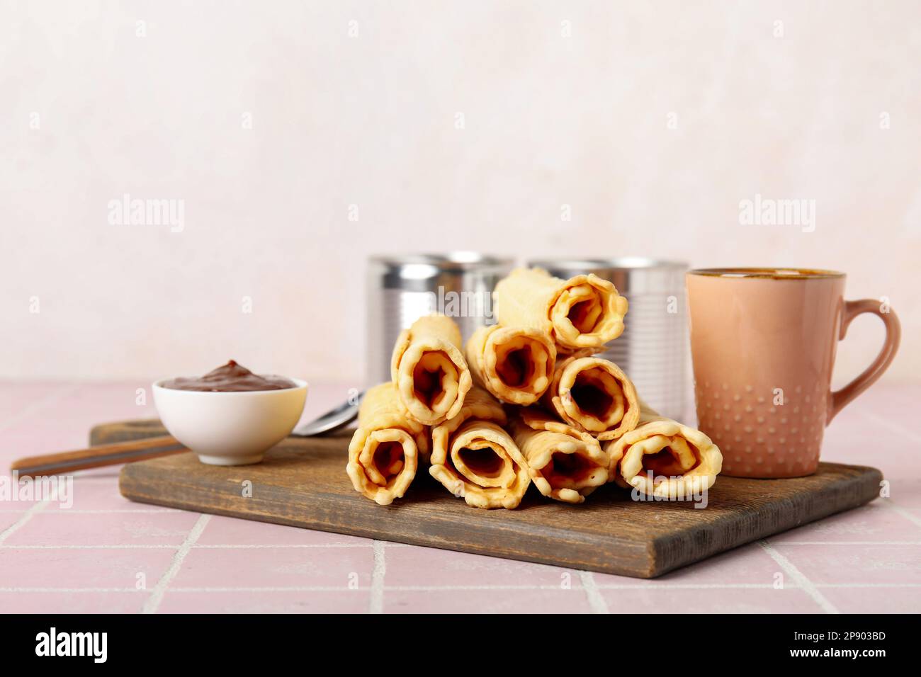 Board with stack of delicious wafer rolls and cup of coffee on pink ...