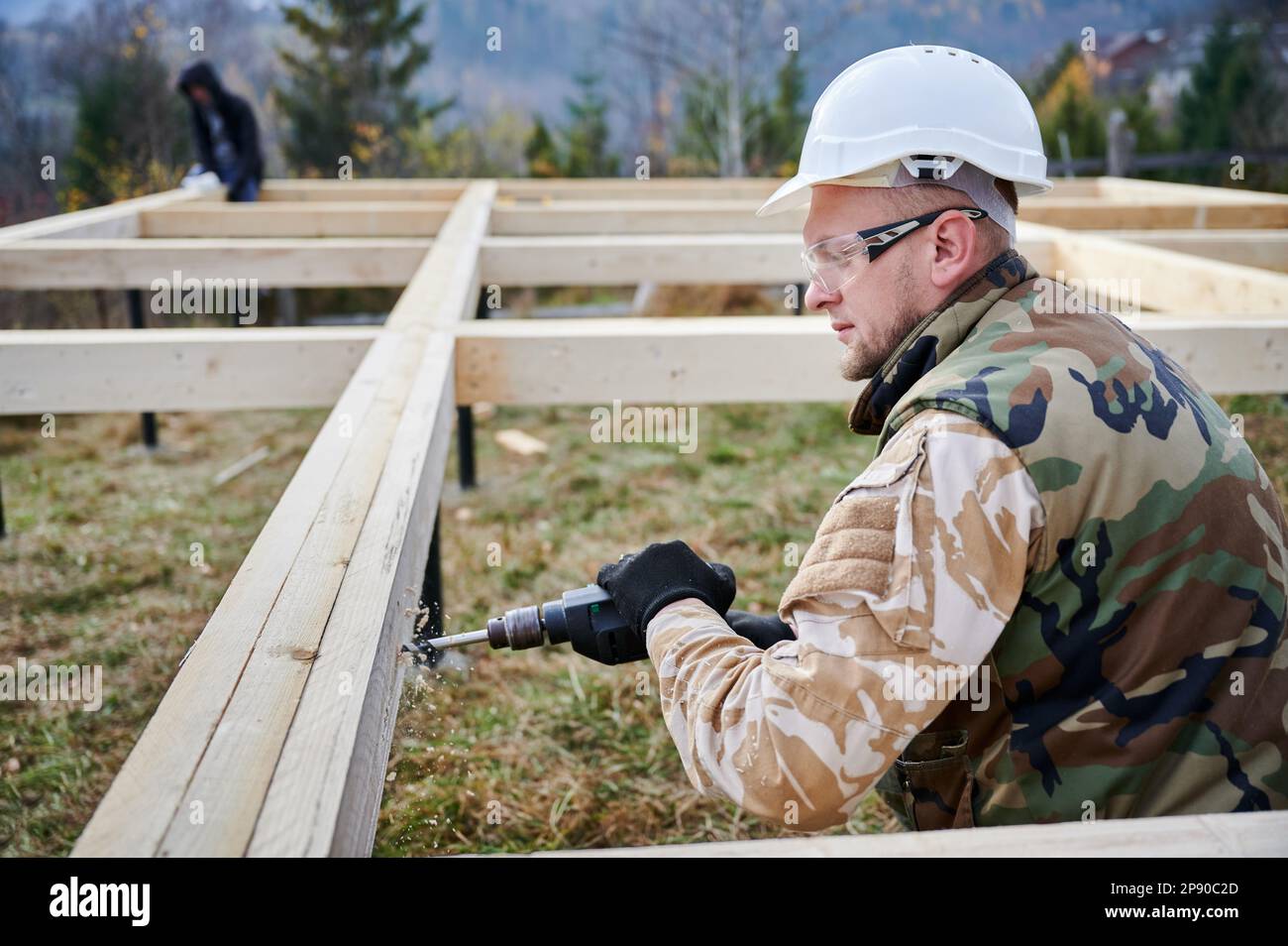 Man building wooden frame house on pile foundation. Male worker drills ...