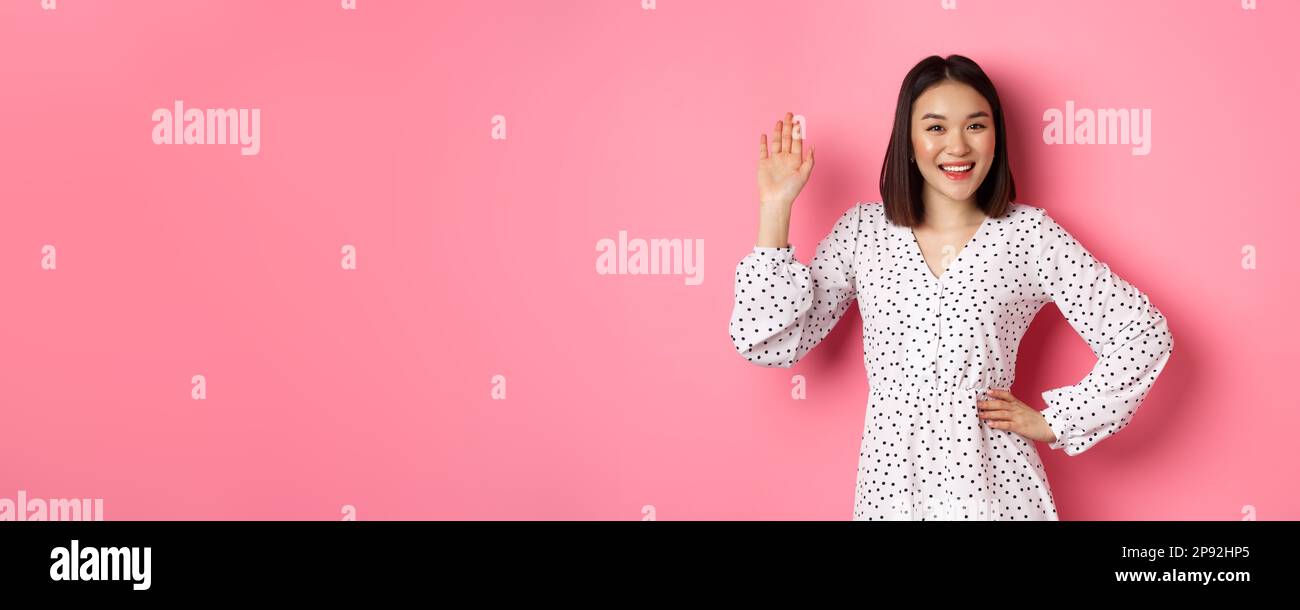 Beautiful asian woman in dress saying hello, waving hand to greet and ...