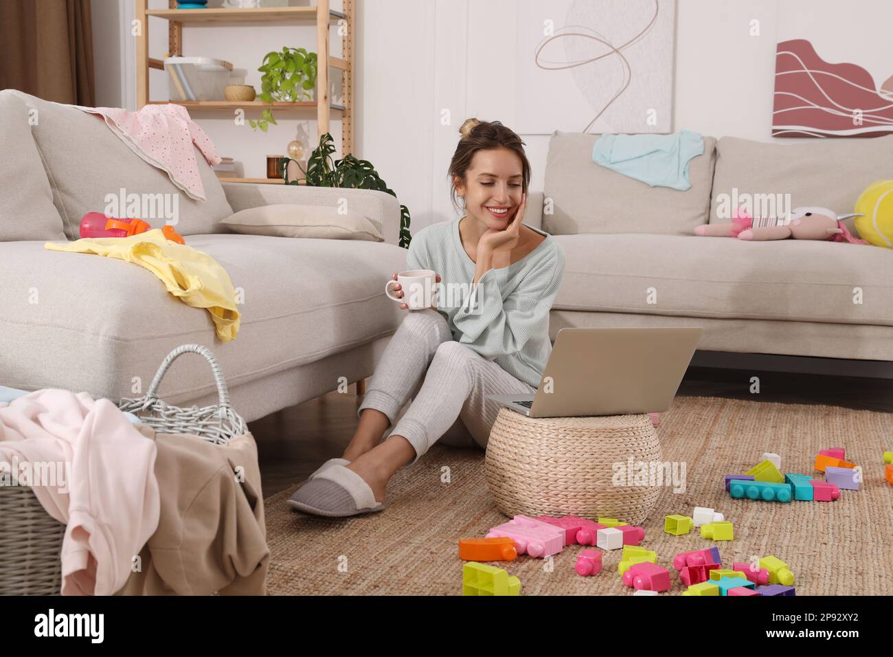 Young mother drinking tea and working with laptop on floor in messy ...