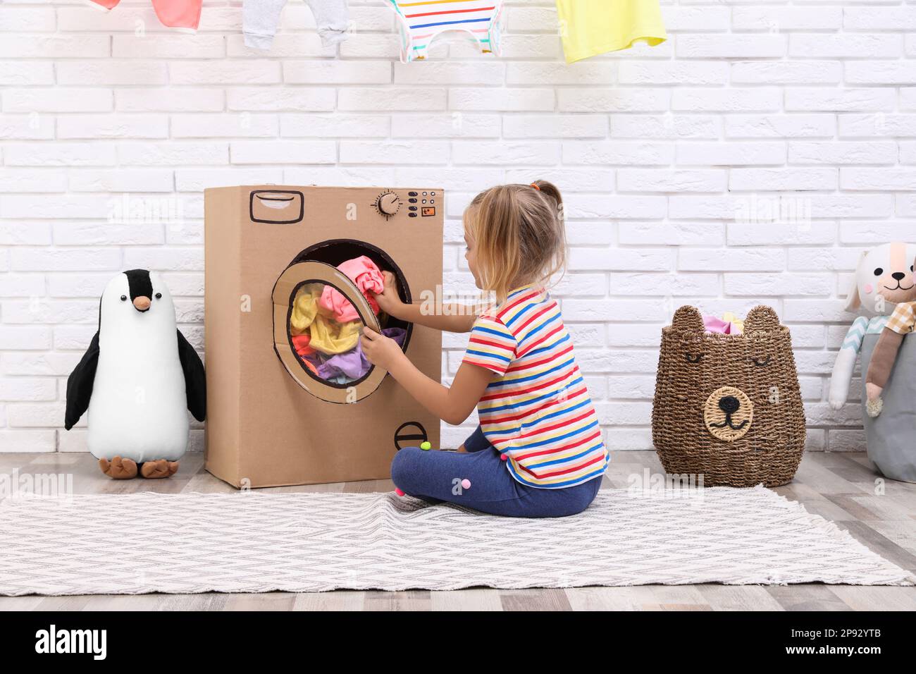 Little girl putting laundry into toy cardboard washing machine indoors ...