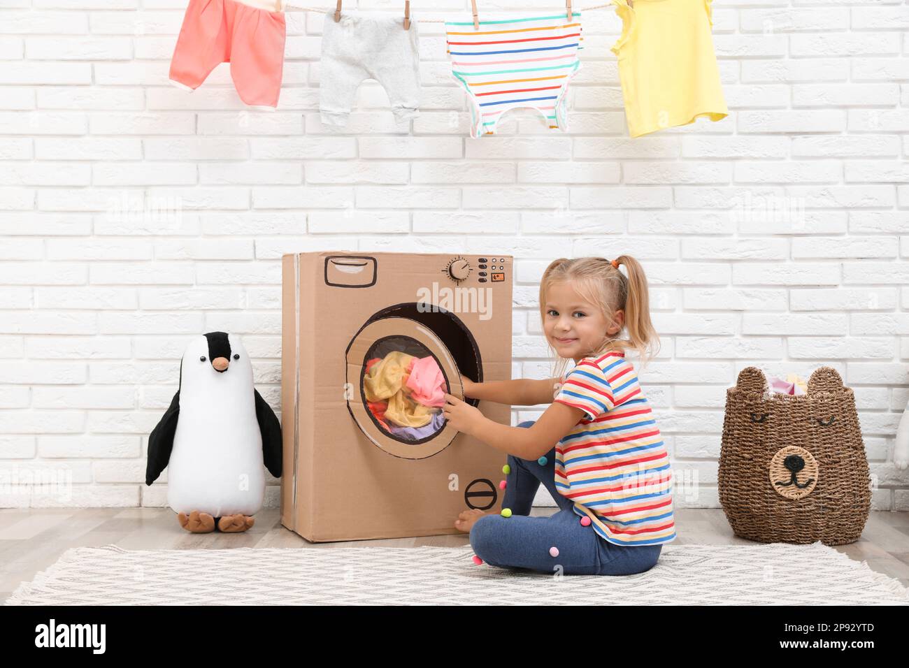 Little girl putting laundry into toy cardboard washing machine indoors ...