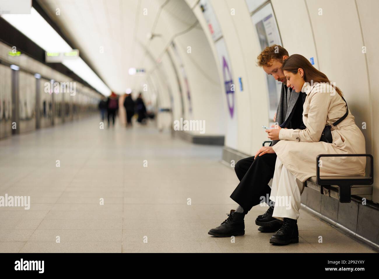 Couple is sitting on a subway station bench and watching something on a ...
