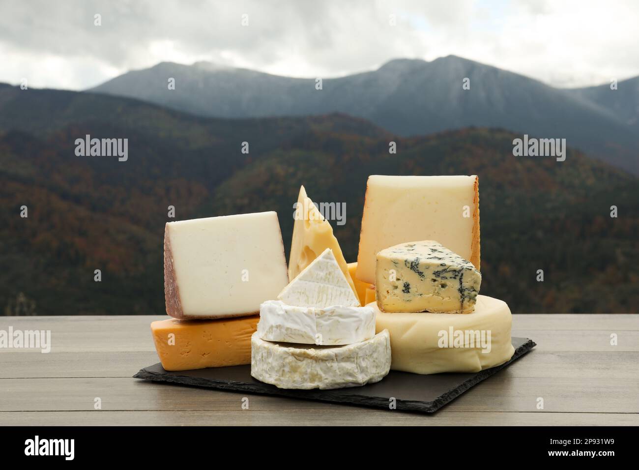 Different types of delicious cheeses on wooden table against mountain ...