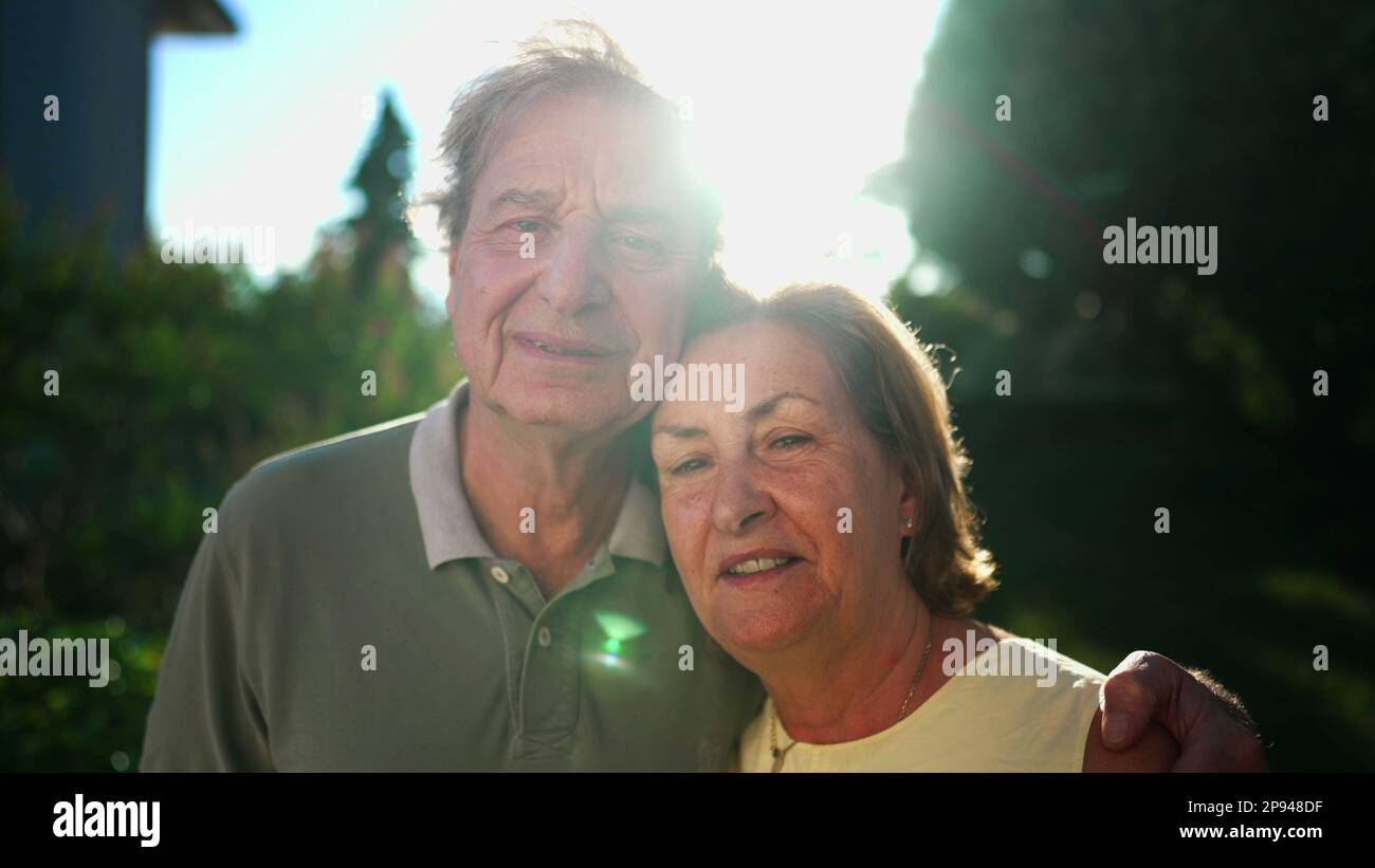 A happy married older couple looking at camera at park with sunlight ...