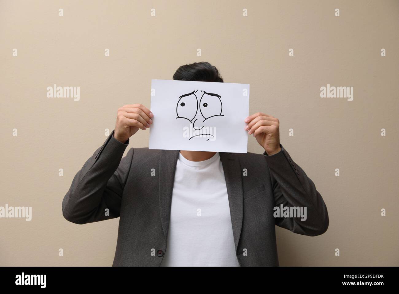 Man hiding emotions using card with drawn frowning face on beige ...