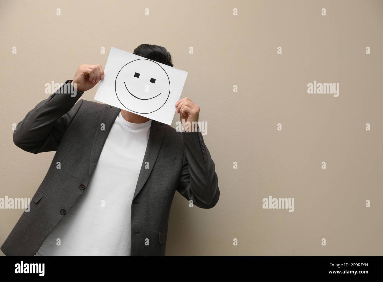Man hiding emotions using card with drawn smiling face on beige ...