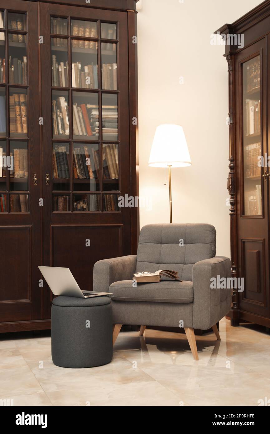 Comfortable armchair with pouf, laptop and lamp near wooden bookcase in ...
