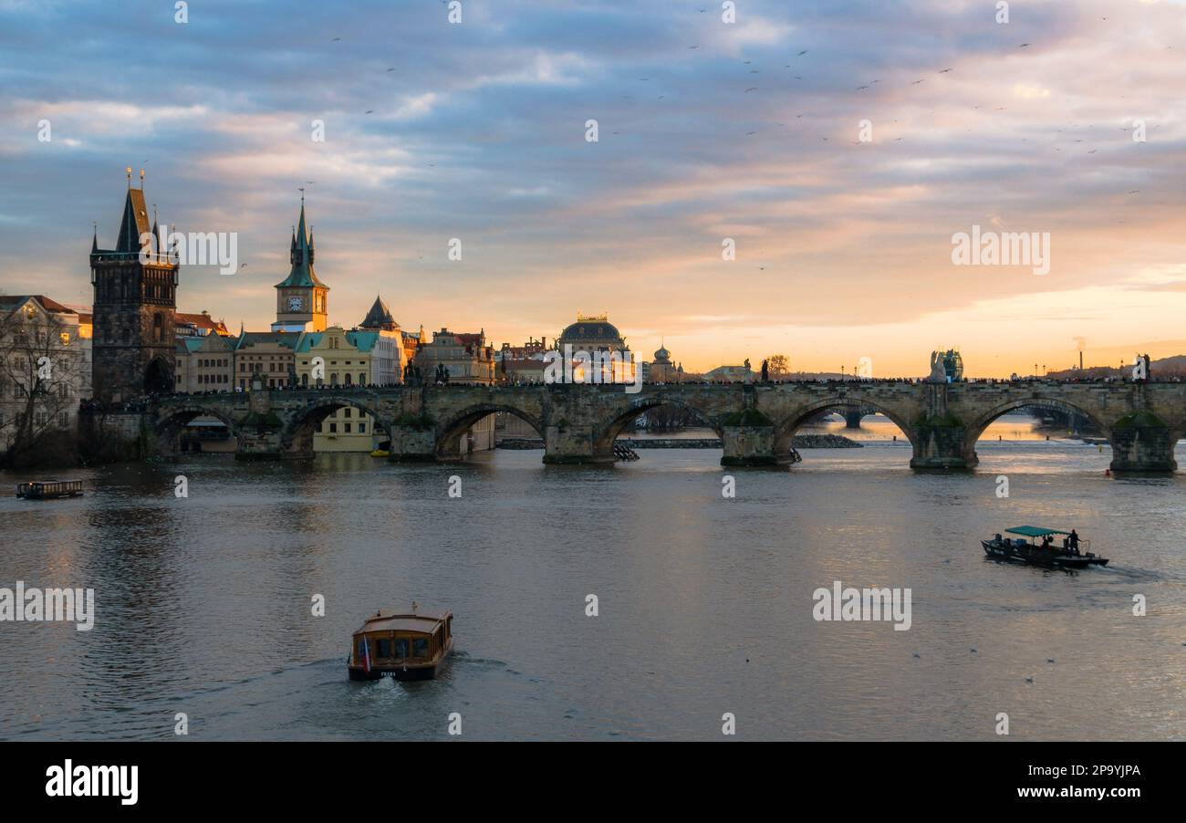 Sunset view over Charles Bridge in Prague, pedestrians waling on the bridge, boat cruises and ...