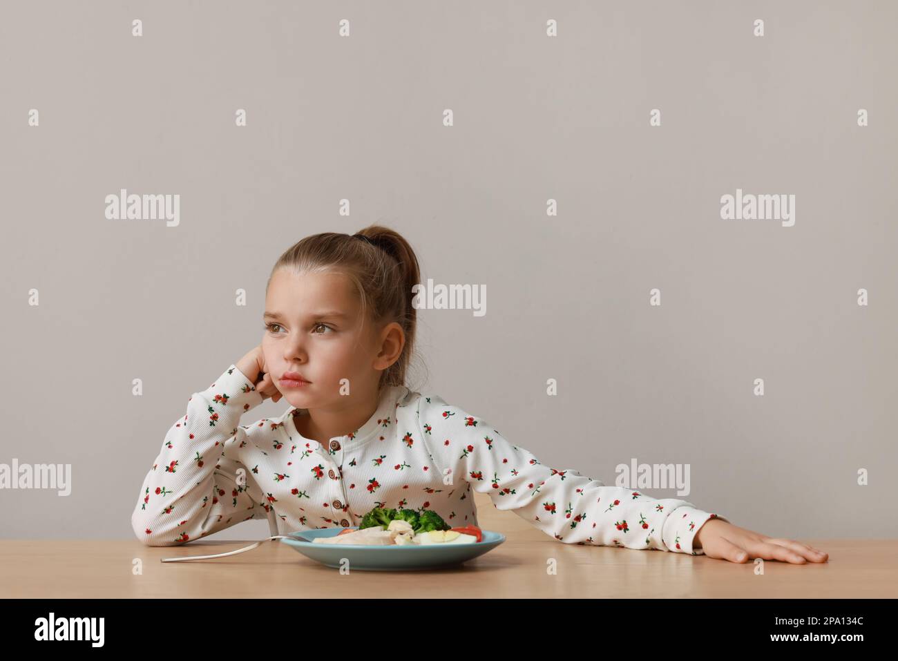 Cute little girl refusing to eat her dinner at table on grey background ...