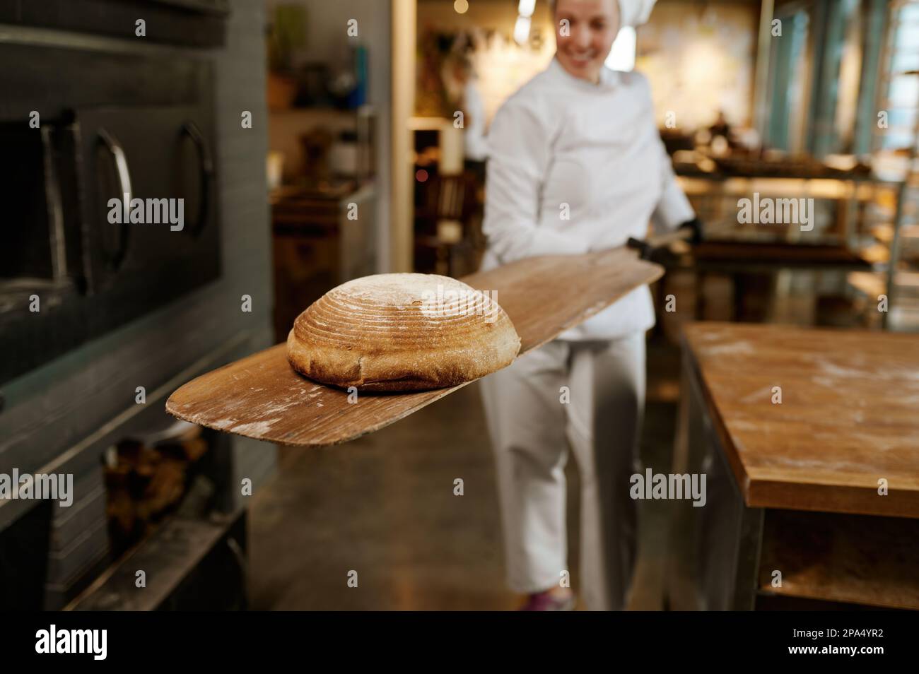 Young bakery worker using wooden shovel to take bread from stove oven ...
