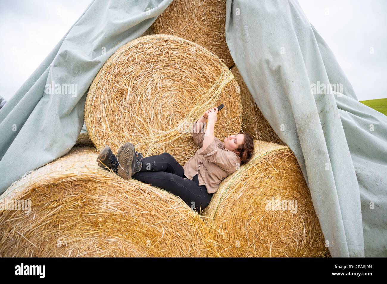 Young woman with brown curly hair is lying on a stack of hay balls and ...