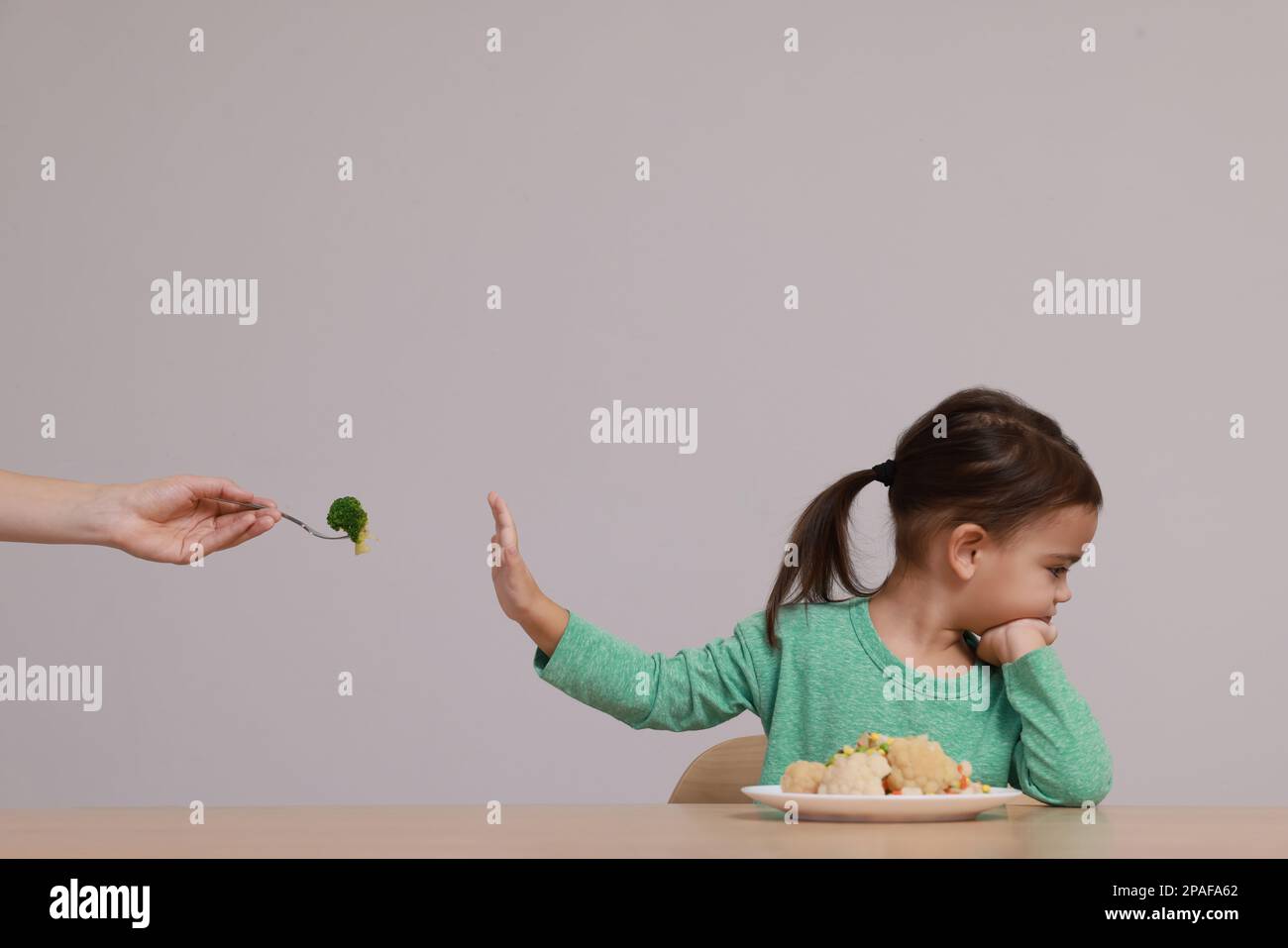 Cute little girl refusing to eat vegetables at table on grey background ...