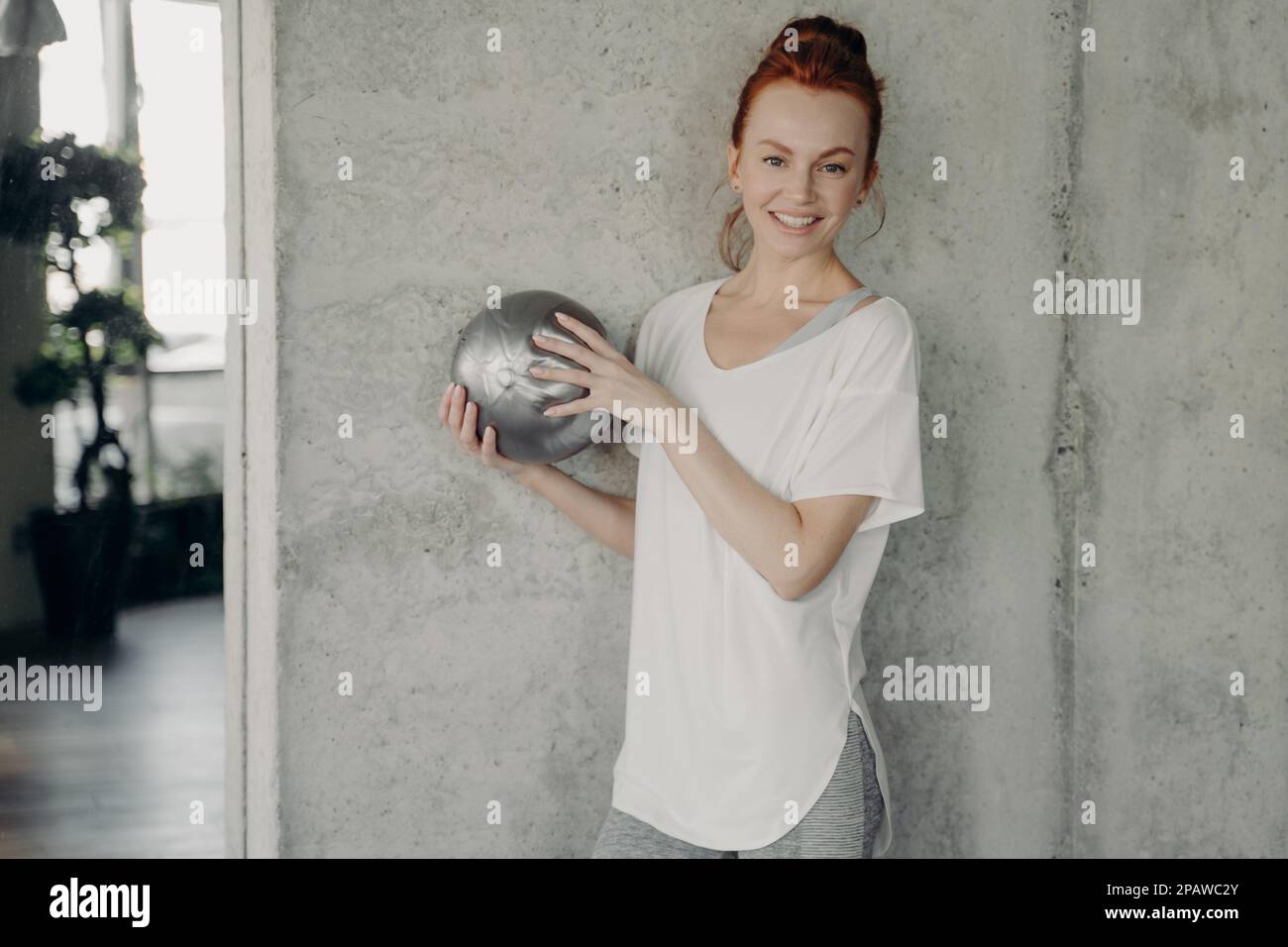Positive slim young female with foxy hair stands by wall in white sport ...