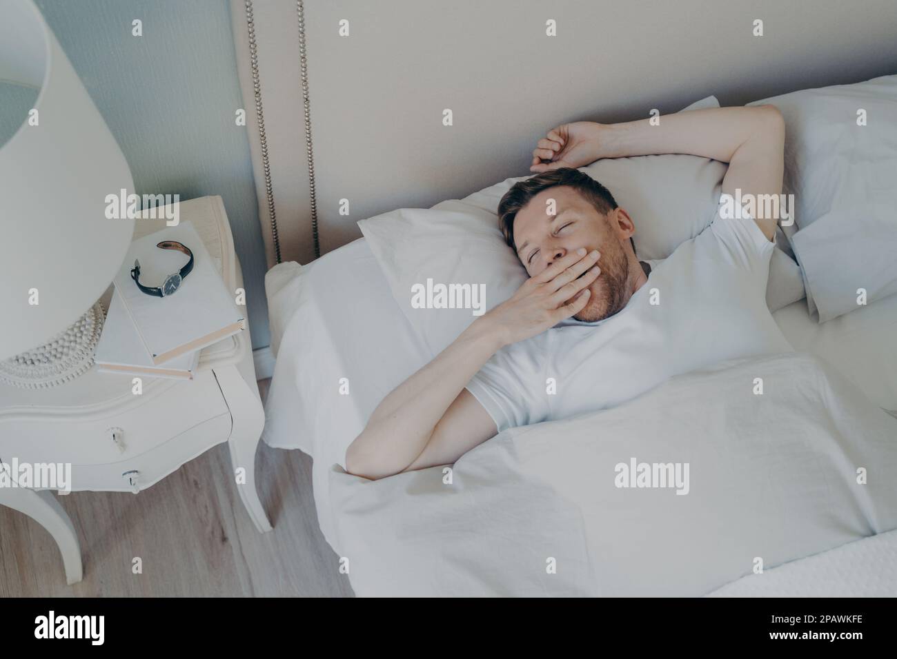 Young sleepy man waking up in morning after night sleep while lying in ...