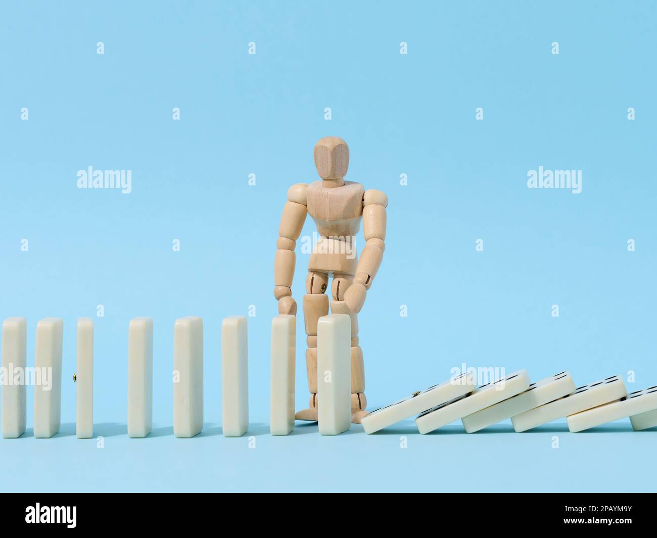 A wooden puppet toy holds back a falling domino on a blue background ...