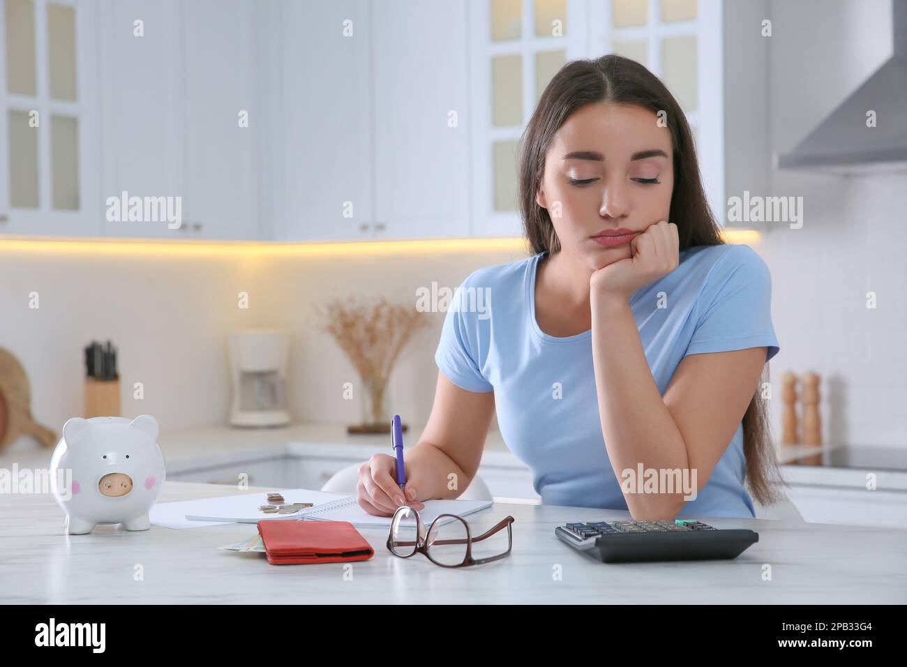Sad young woman counting money with calculator at table in kitchen ...