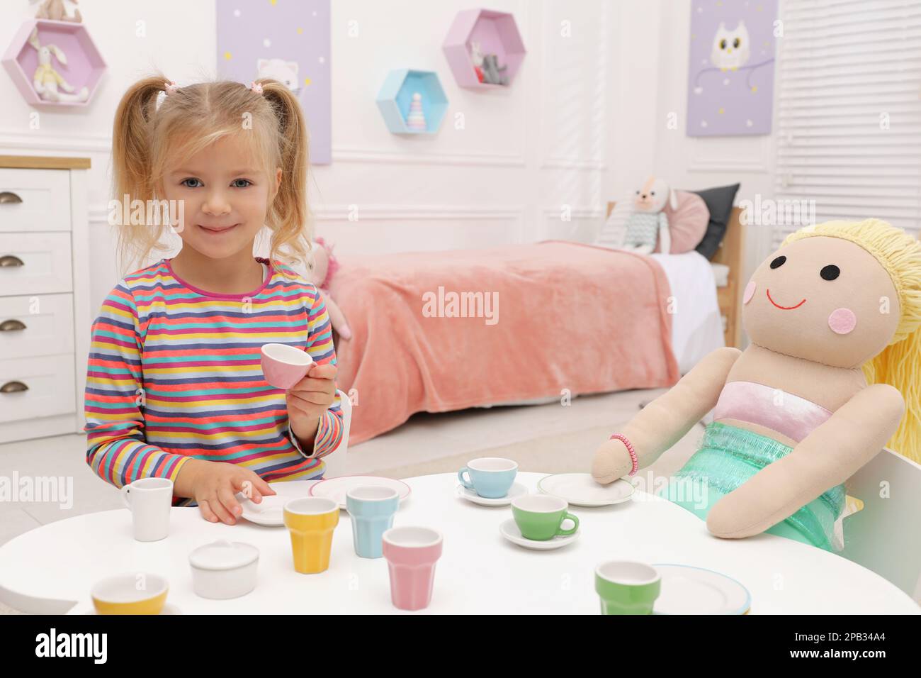 Cute little girl playing tea party with doll at table in room, space ...