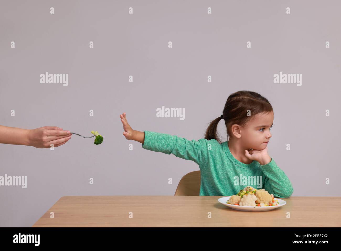 Cute little girl refusing to eat vegetables at table on grey background ...