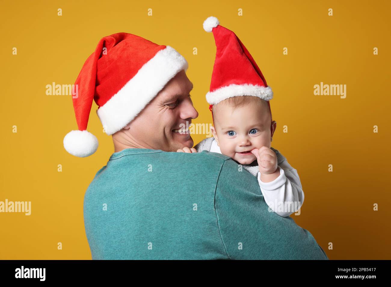 Happy father with cute baby wearing Santa hats on yellow background ...