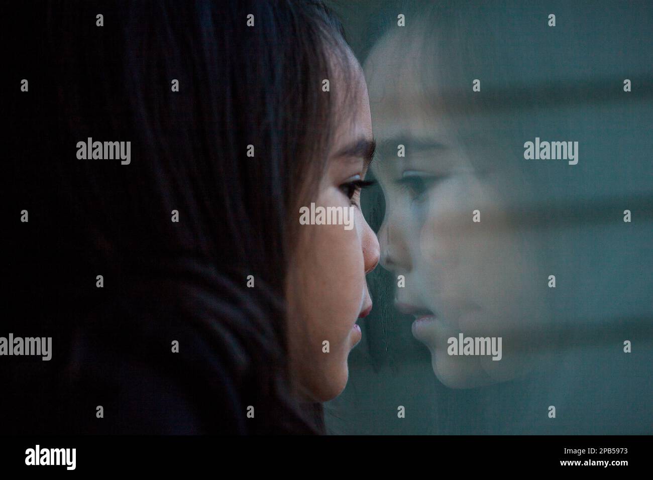 A young girl's face is reflected in the window through which she's ...