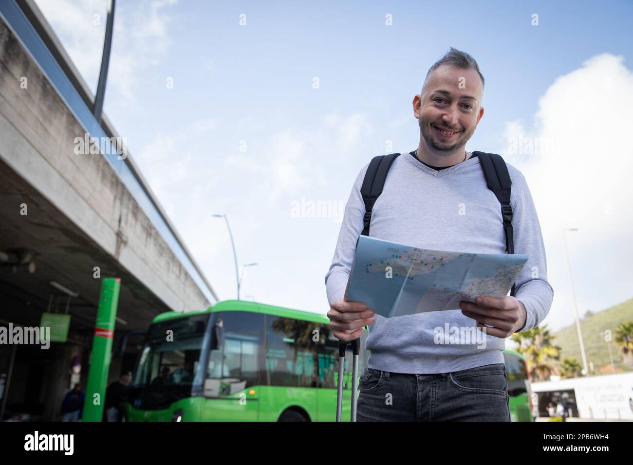A smiling traveler holding a map and wearing a backpack at a bus ...