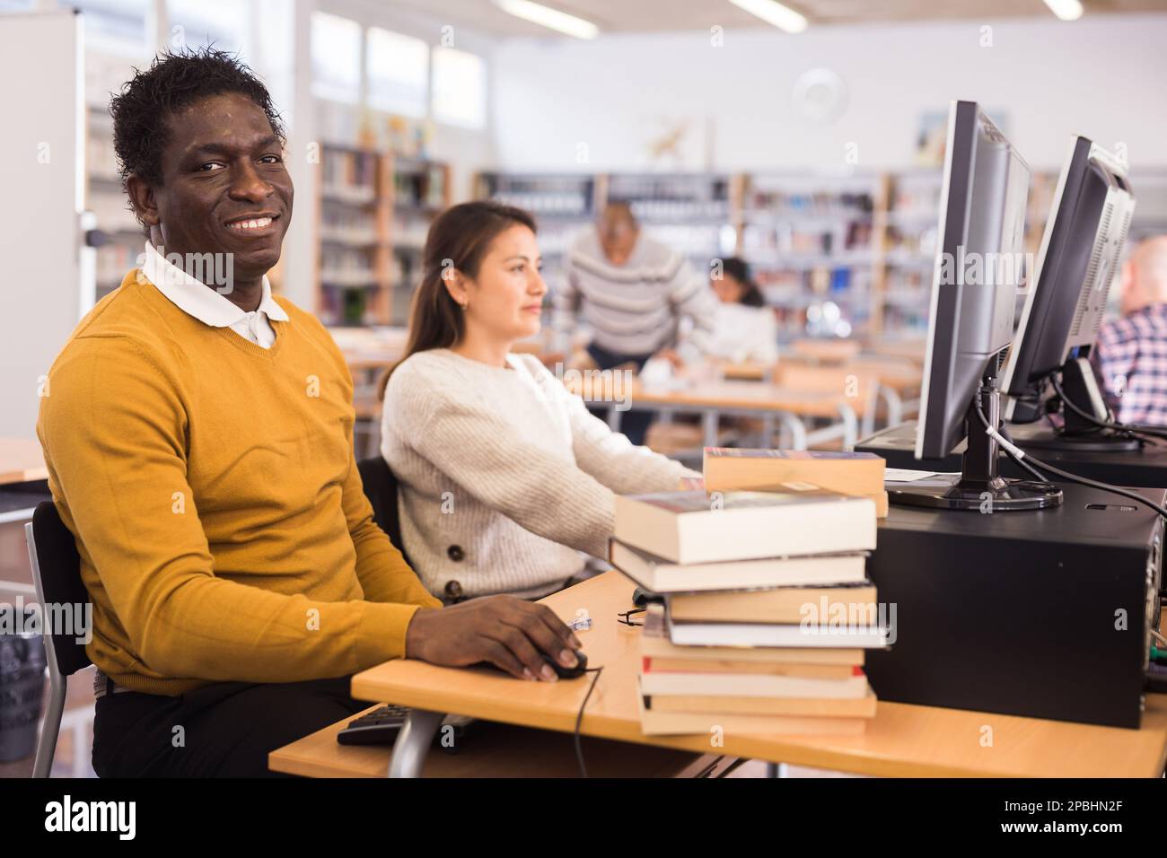 Cheerful aframerican man reading books and using pc in library Stock ...