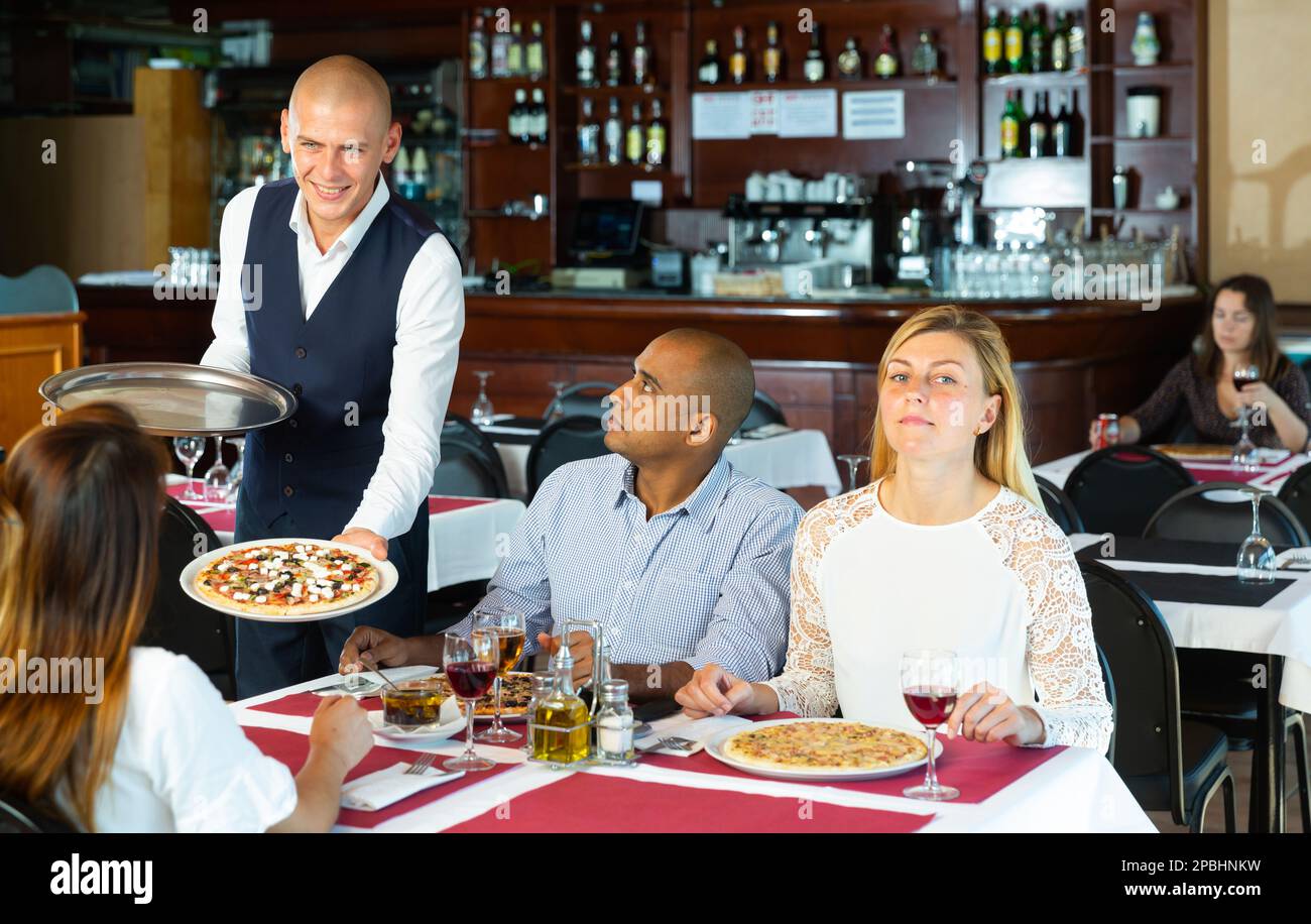 Polite waiter serving pizza to friendly company in pizzeria Stock Photo ...