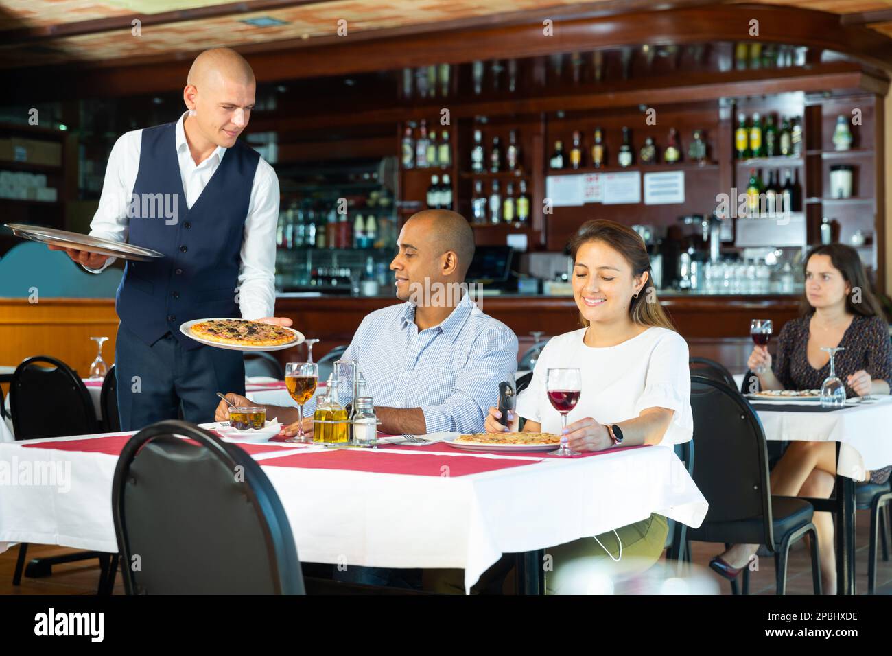 Polite waiter serving pizza to hispanic couple in restaurant Stock ...