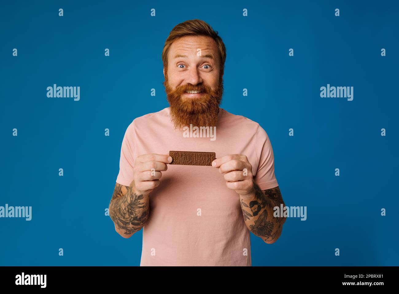 White hungry bearded man eating chocolate bar isolated on blue ...