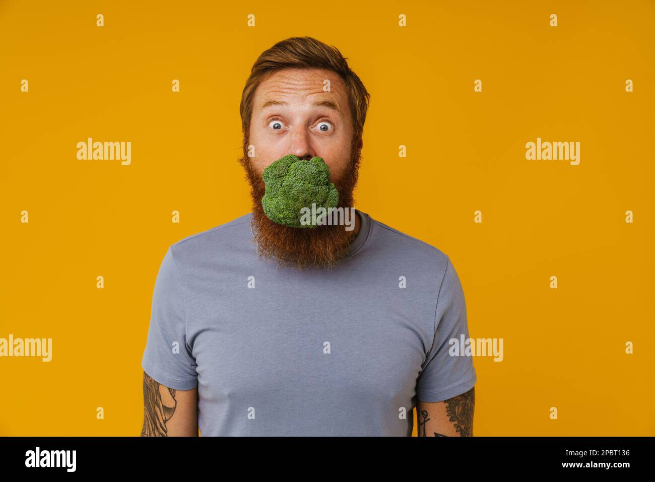 Bearded happy man eating broccoli while standing isolated over yellow ...