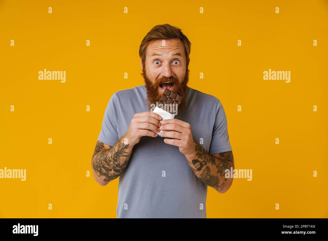 Bearded happy man eating chocolate bar while standing isolated over ...