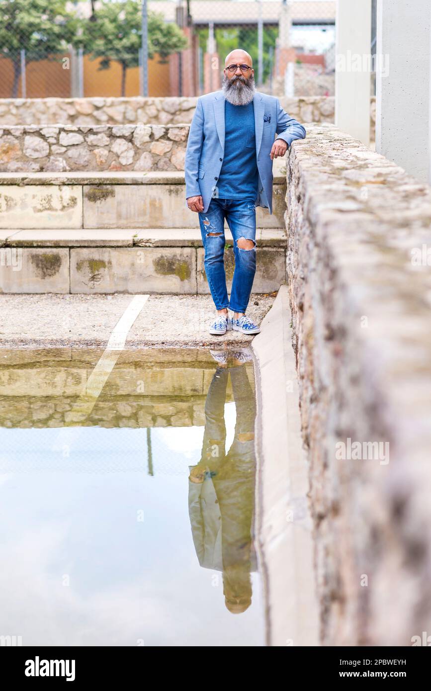 bald, bearded man standing leaning against a park wall by the water ...