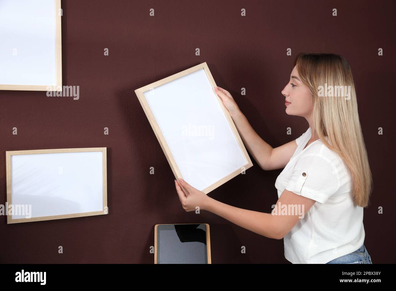Young woman hanging empty frame on brown wall indoors. Mockup for ...