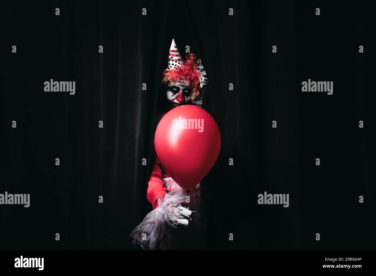 Terrifying clown with red air balloon hiding behind black curtains ...