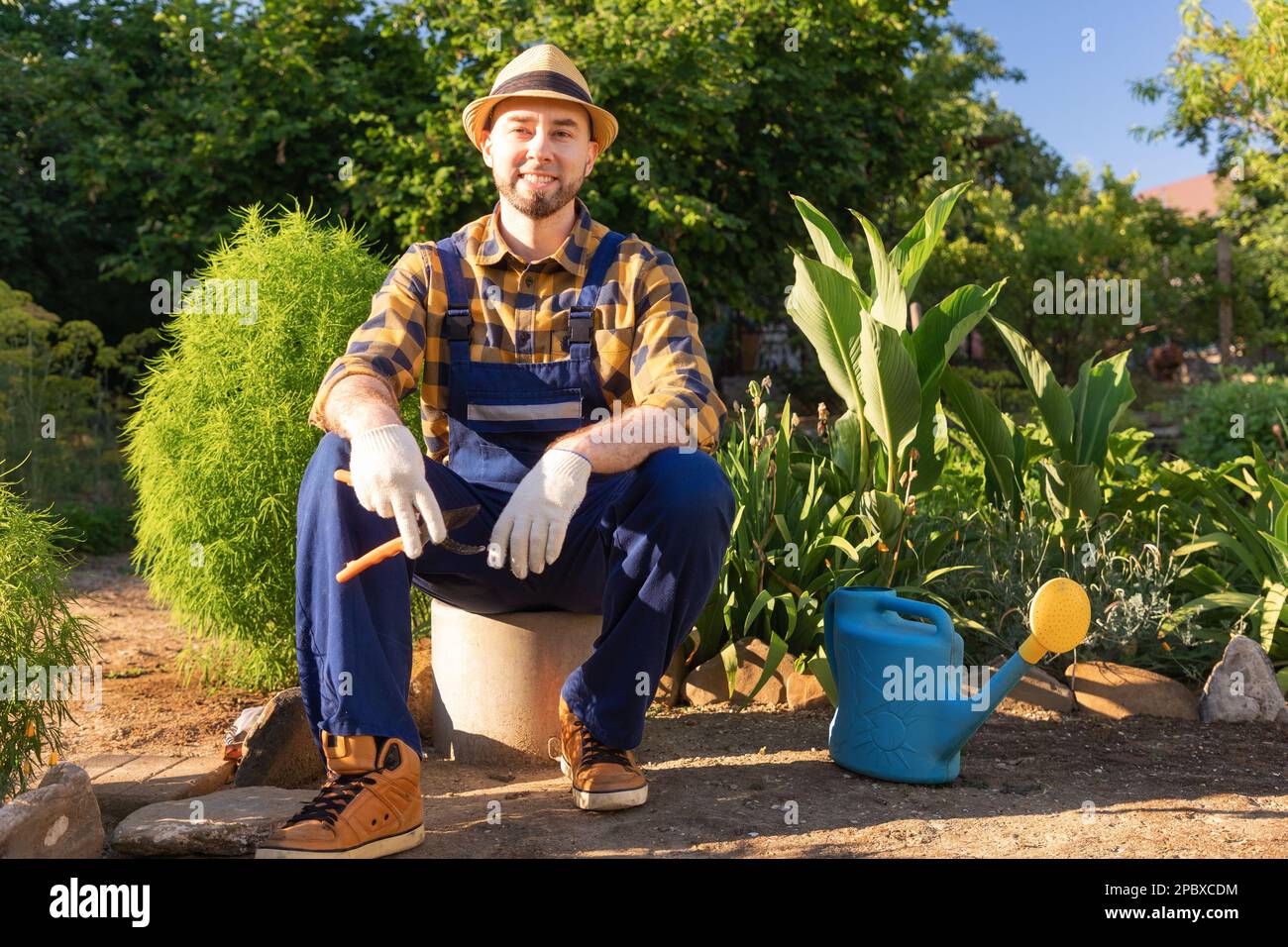 Happy bearded, handsome man in overalls and a straw hat sits and relax ...