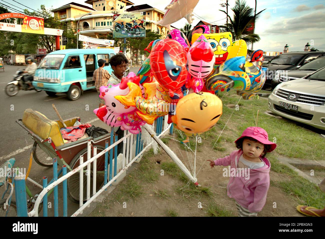 A child is standing by a mobile balloon vendor in a background of road ...