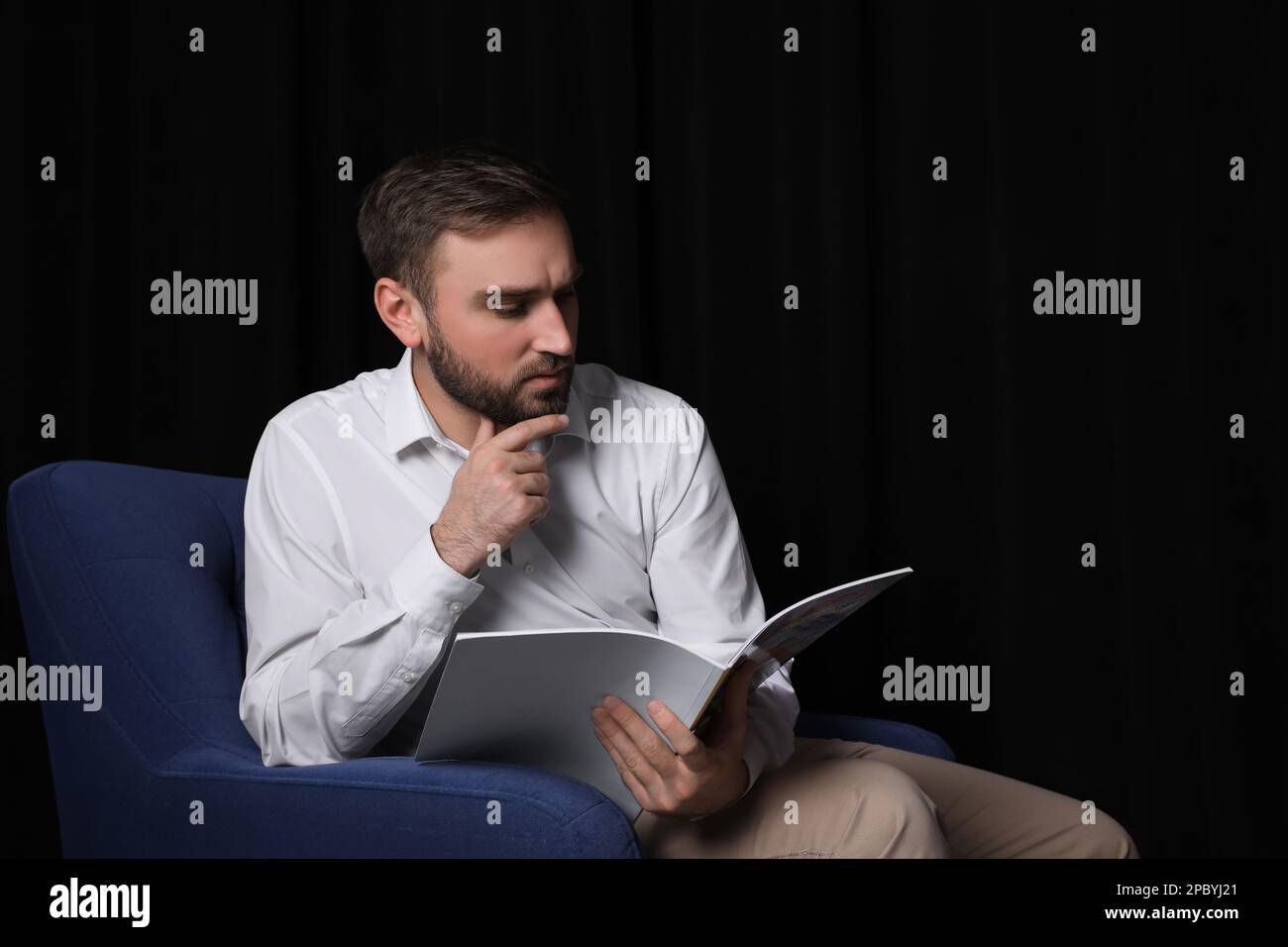 Handsome man reading magazine in armchair against dark background Stock ...