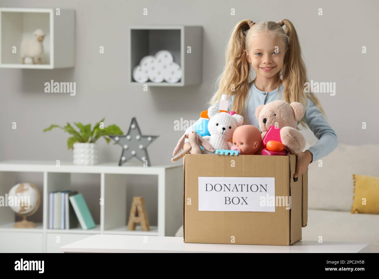 Cute little girl holding donation box with toys at home, space for text ...